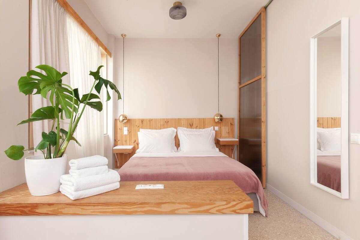 Quarto de hotel moderno com cabeceira de madeira, uma grande planta em vaso, toalhas brancas dobradas com cuidado e mesa de cabeceira de madeira.