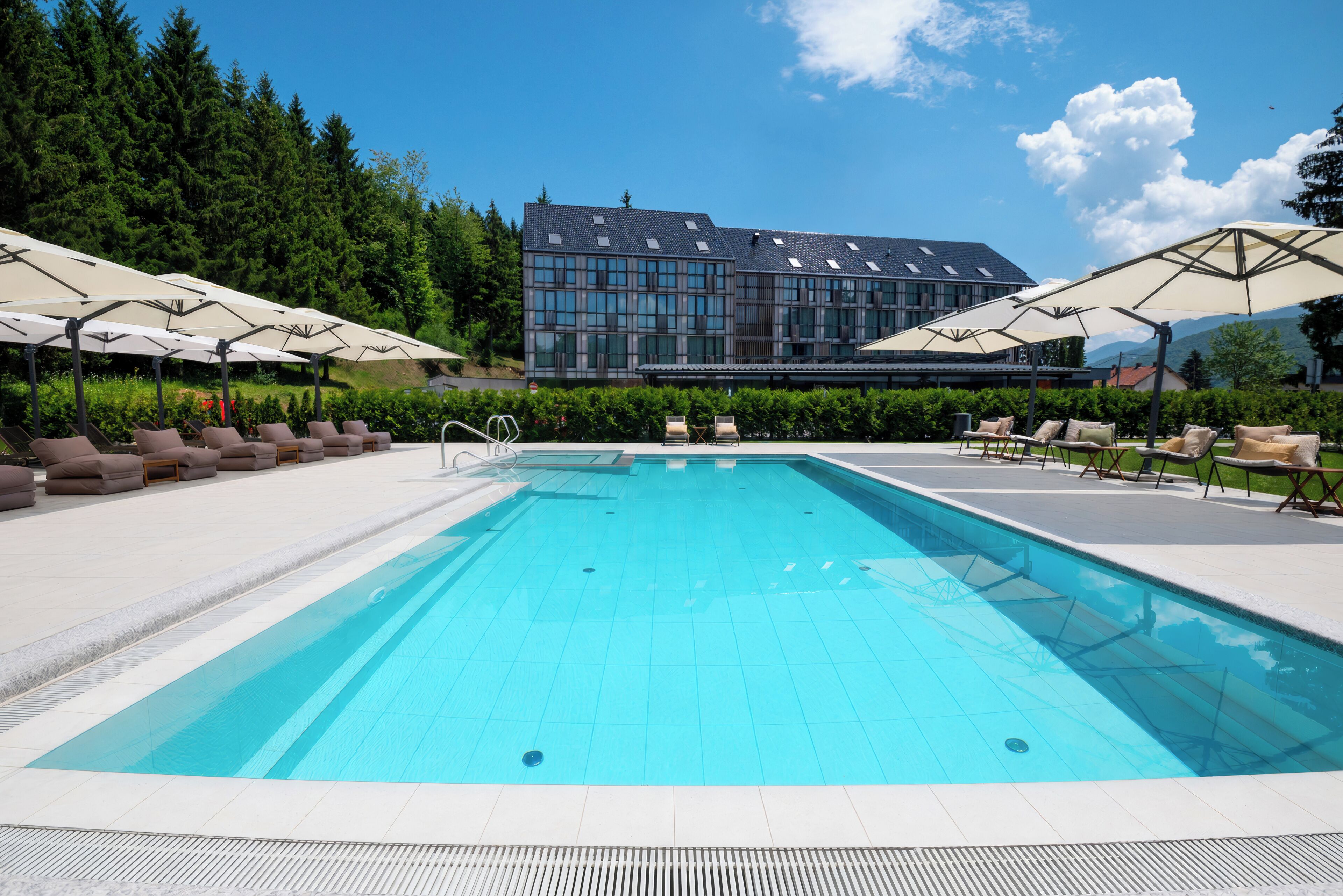 Piscina com espreguiçadeiras e guarda-sóis, cercada por um edifício com grandes janelas e vista para árvores e montanhas.