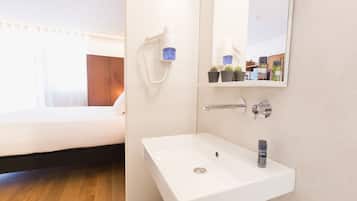 Quarto de hotel com cama, pia branca, espelho e um pequeno estante com produtos de higiene pessoal.
