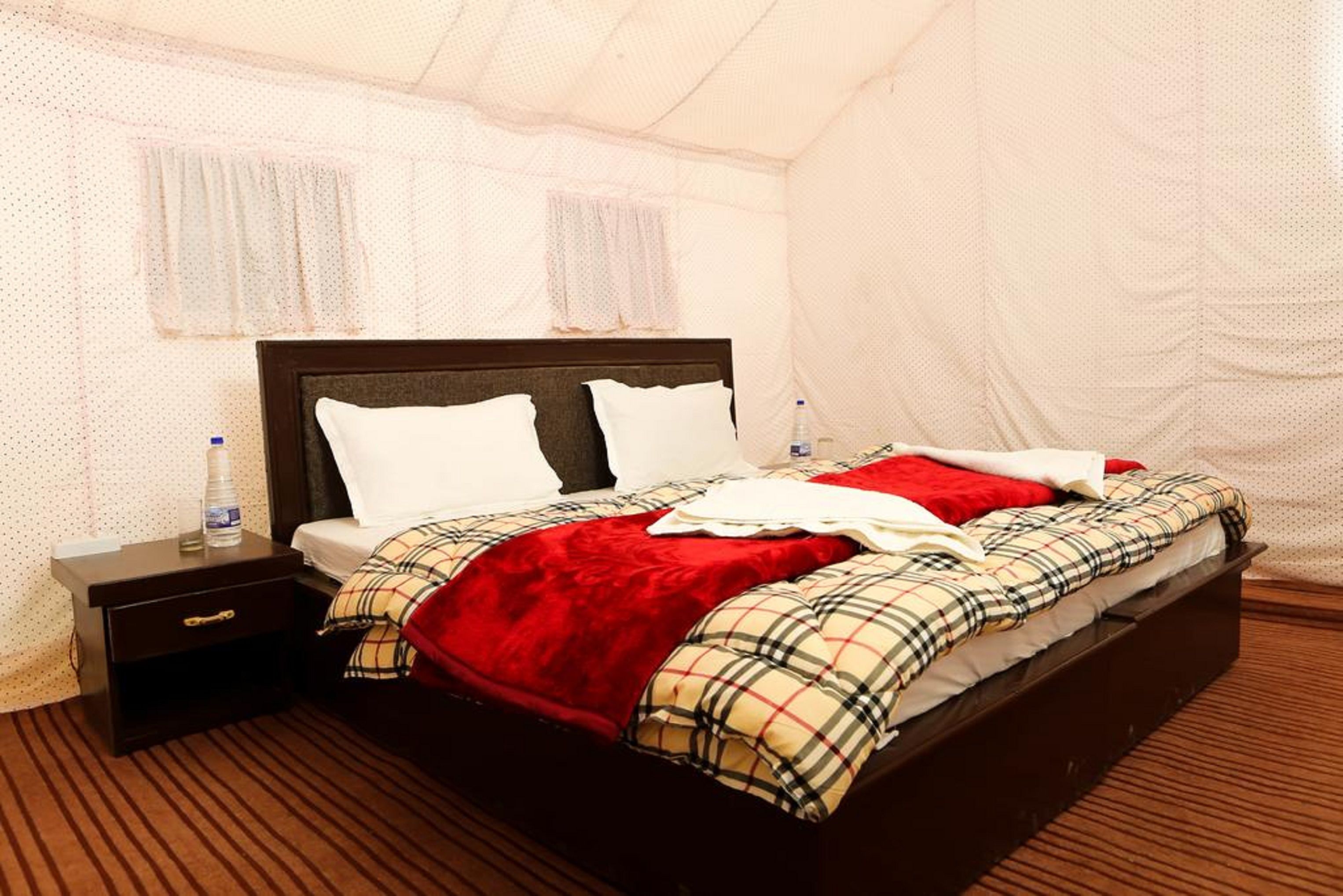 Uma cama bem arrumada com manta xadrez e almofadas, um criado-mudo de madeira com uma garrafa e uma janela com cortinas leves.