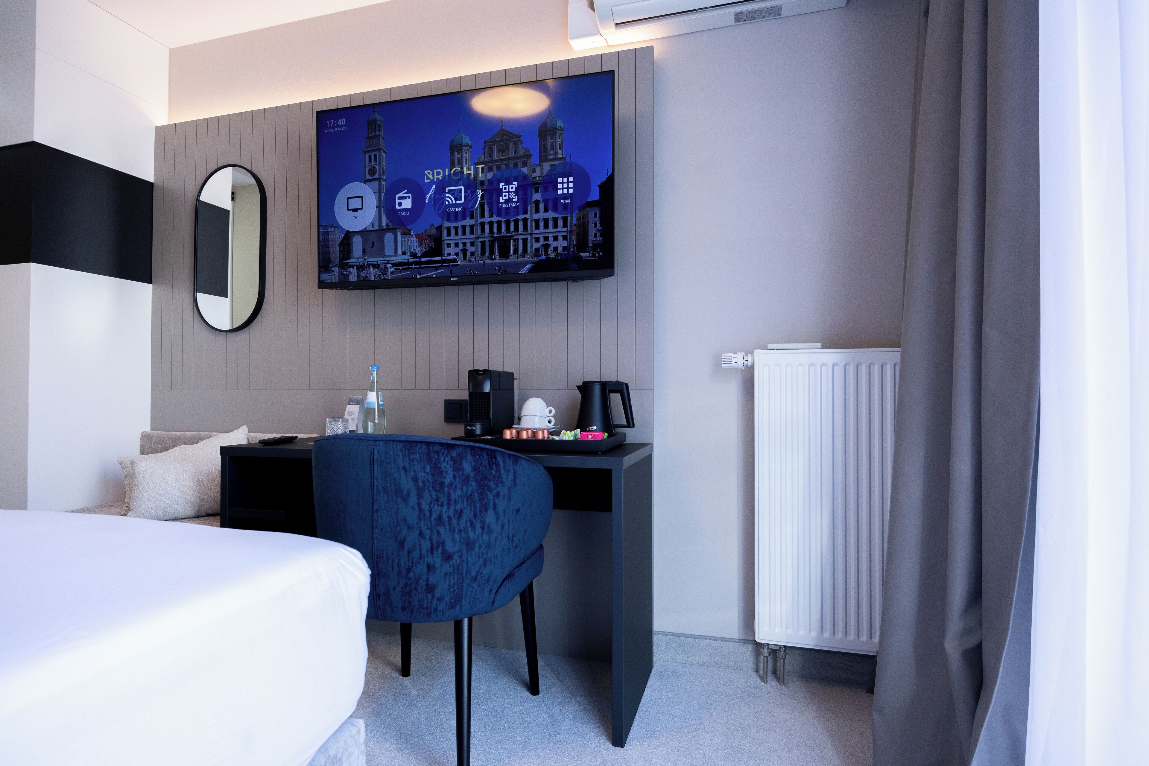 Quarto de hotel moderno com cama, escrivaninha com cadeira, televisão na parede e radiador.