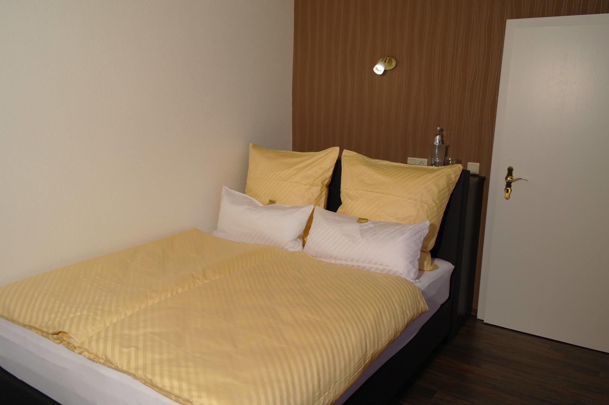 Uma cama bem arrumada com colcha amarela e travesseiros brancos, uma cabeceira de madeira e uma porta com puxador de latão.