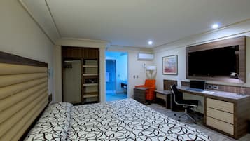 Quarto de hotel com cama, escrivaninha, cadeira, televisão e guarda-roupa.