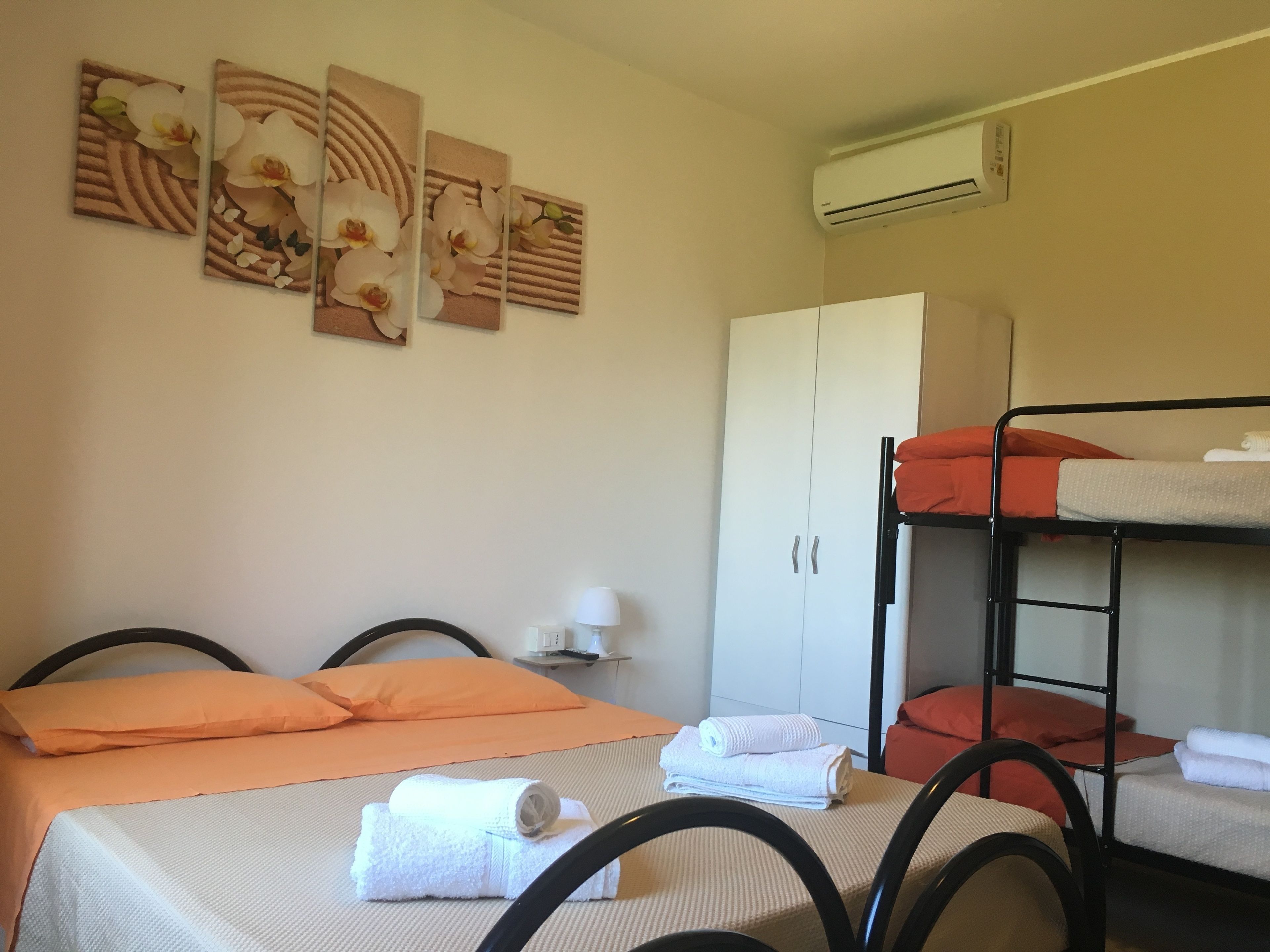 Quarto de hotel com uma cama beliche, uma cama de solteiro com roupa de cama laranja, uma toalha branca na cama de solteiro, um armário branco e arte nas paredes com flores.