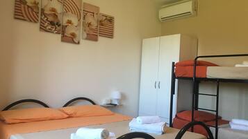 Quarto de hotel com uma cama beliche, uma cama de solteiro com roupa de cama laranja, uma toalha branca na cama de solteiro, um armário branco e arte nas paredes com flores.