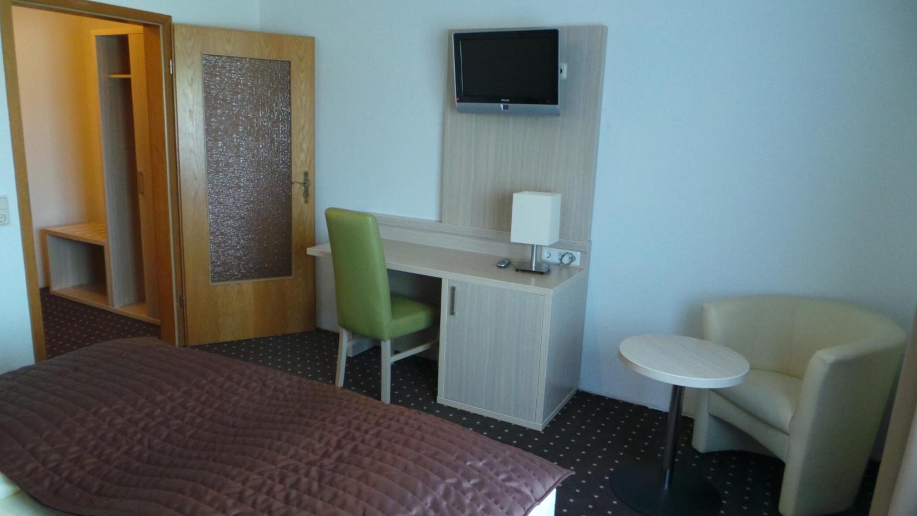 Quarto de hotel com cama, escrivaninha com cadeira, televisão na parede e uma pequena mesa redonda.