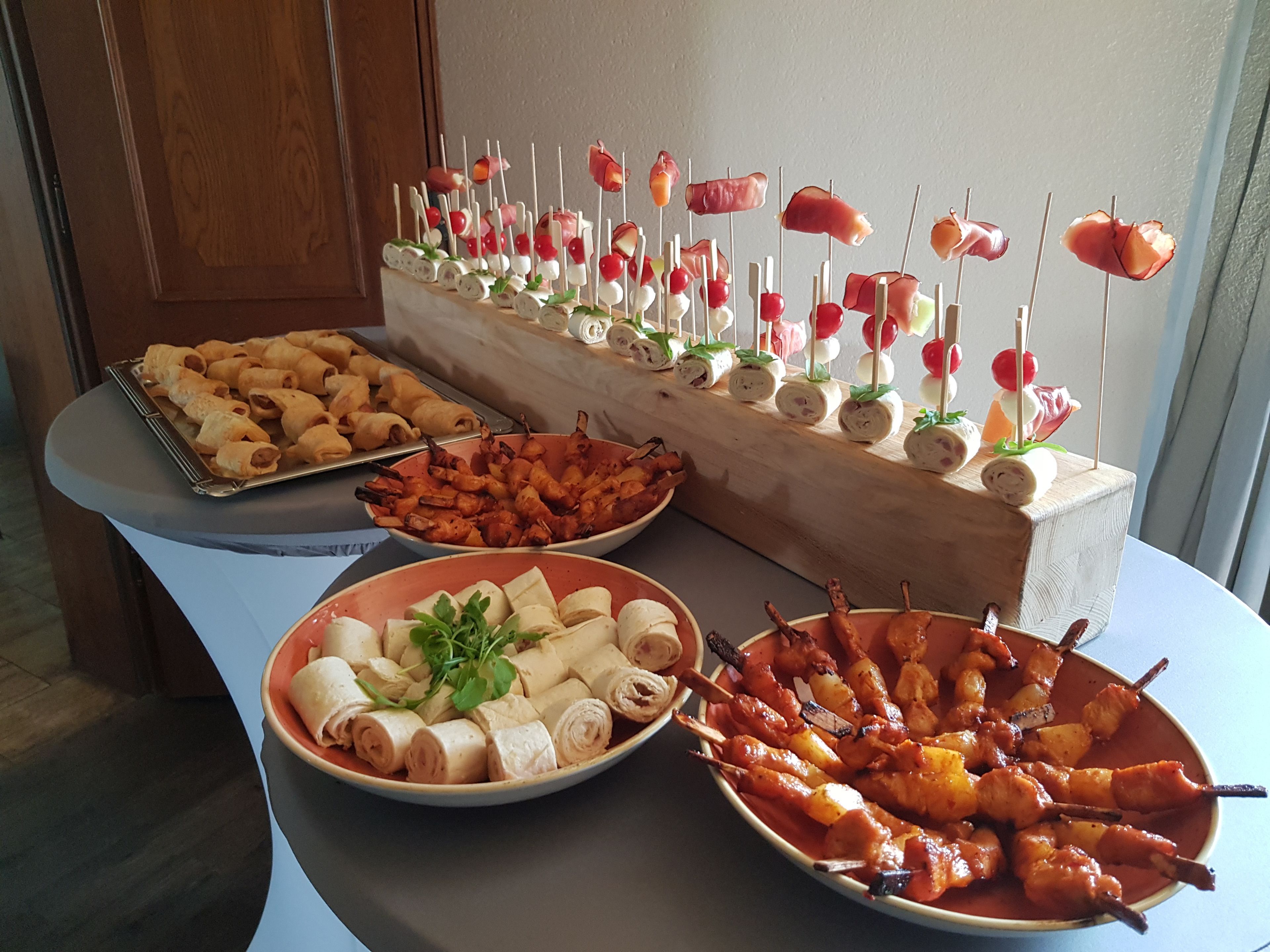 Uma mesa de buffet com diversos alimentos, incluindo carnes enroladas, espetinhos e uma bandeja de doces.