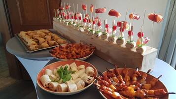 Uma mesa de buffet com diversos alimentos, incluindo carnes enroladas, espetinhos e uma bandeja de doces.