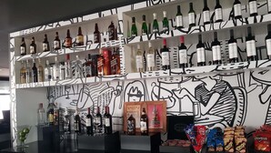 Uma área de bar com prateleiras cheias de garrafas de bebidas, um quadro com cena de bar e petiscos.