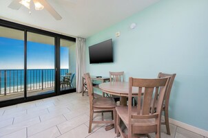 Quarto com varanda vista para a praia, mesa redonda com cadeiras, TV de tela plana na parede e ventilador de teto.