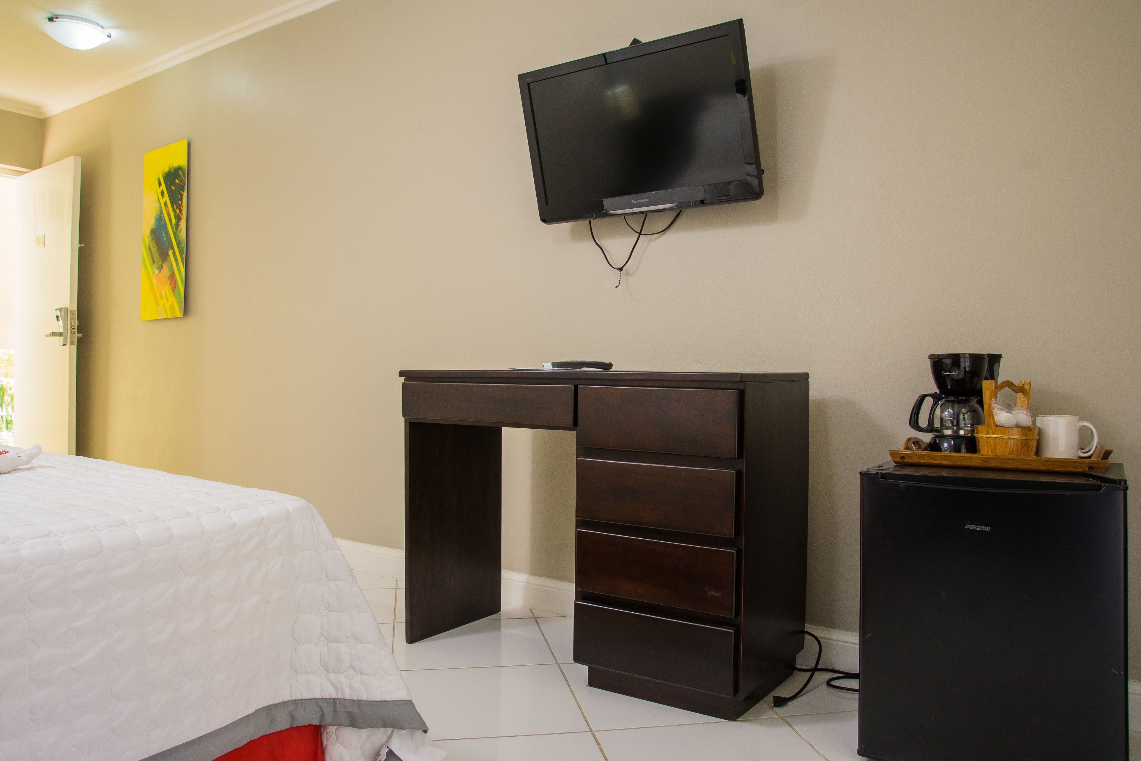 Quarto com cama, escrivaninha com gavetas, televisão na parede e geladeira com cafeteira e caneca em cima.