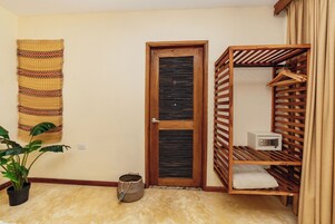 Um quarto com porta de madeira, um detalhe decorativo tecido na parede, uma planta, uma cesta de palha e uma prateleira de madeira com um objeto branco.