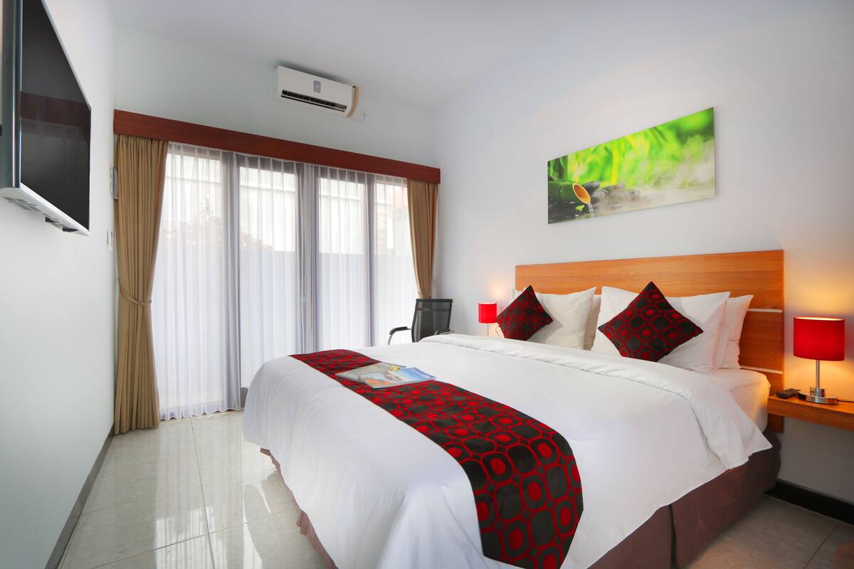 Quarto de hotel com uma cama grande, televisão de tela plana na parede, janela com cortinas finas e um quadro sobre a cama.
