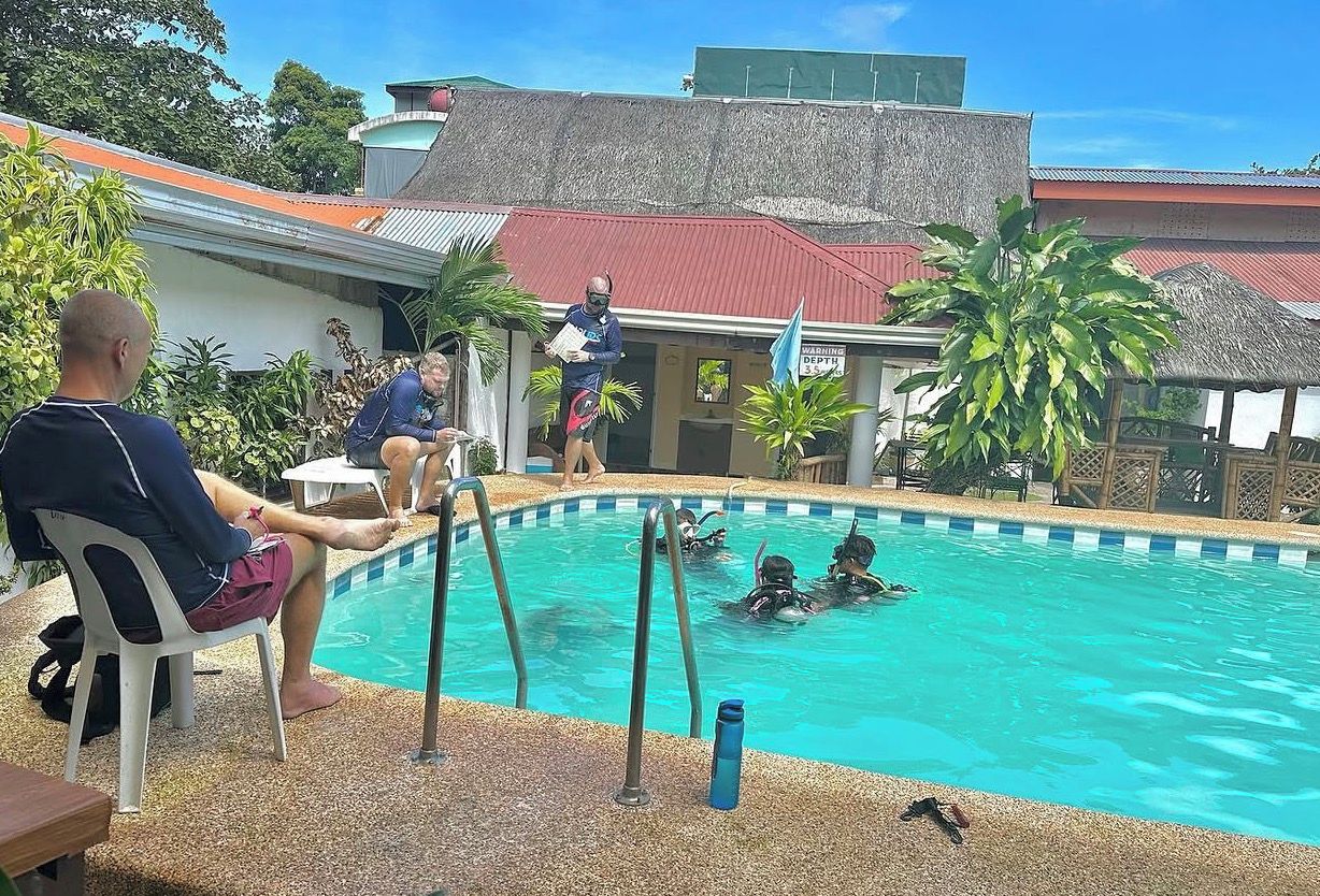 Área de piscina com pessoas e cães, uma bandeira indicando profundidade de 3 metros e um edifício com telhado vermelho ao fundo.