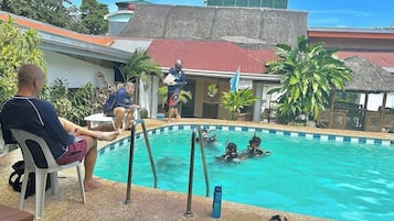 Área de piscina com pessoas e cães, uma bandeira indicando profundidade de 3 metros e um edifício com telhado vermelho ao fundo.