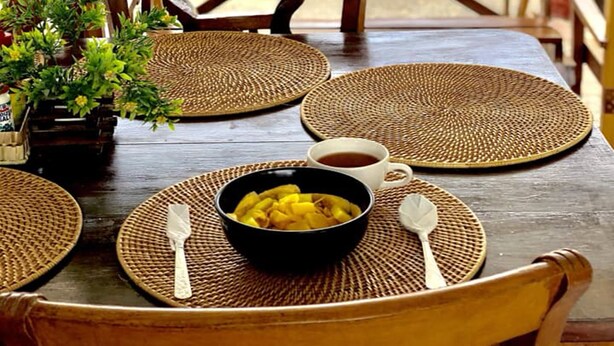 Uma mesa de jantar com um prato de comida, uma chávena de chá e uma colher, com uma piscina e vegetação ao fundo.