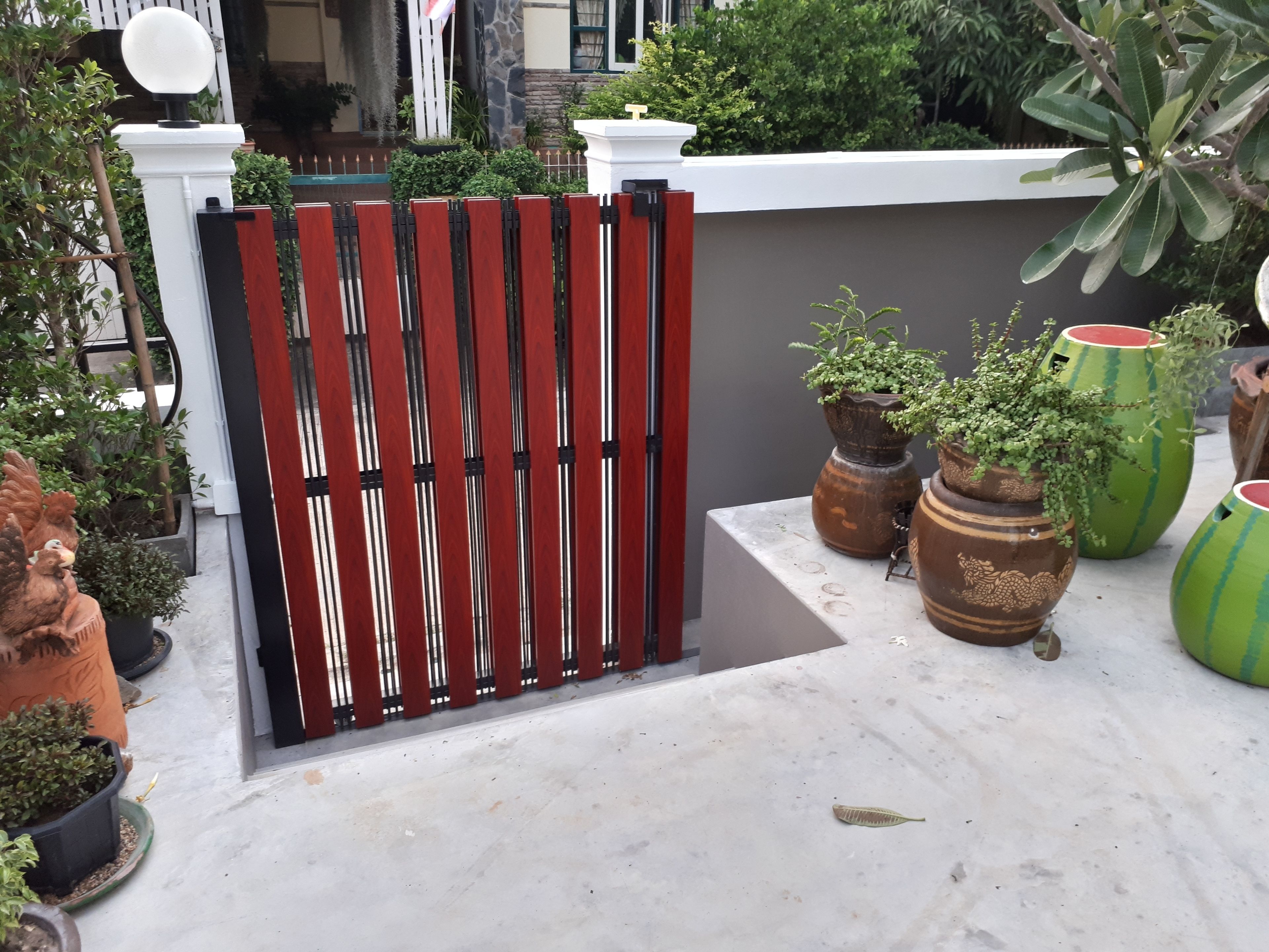 Uma entrada murada com portão vermelho e preto, vasos de plantas e uma parede branca.