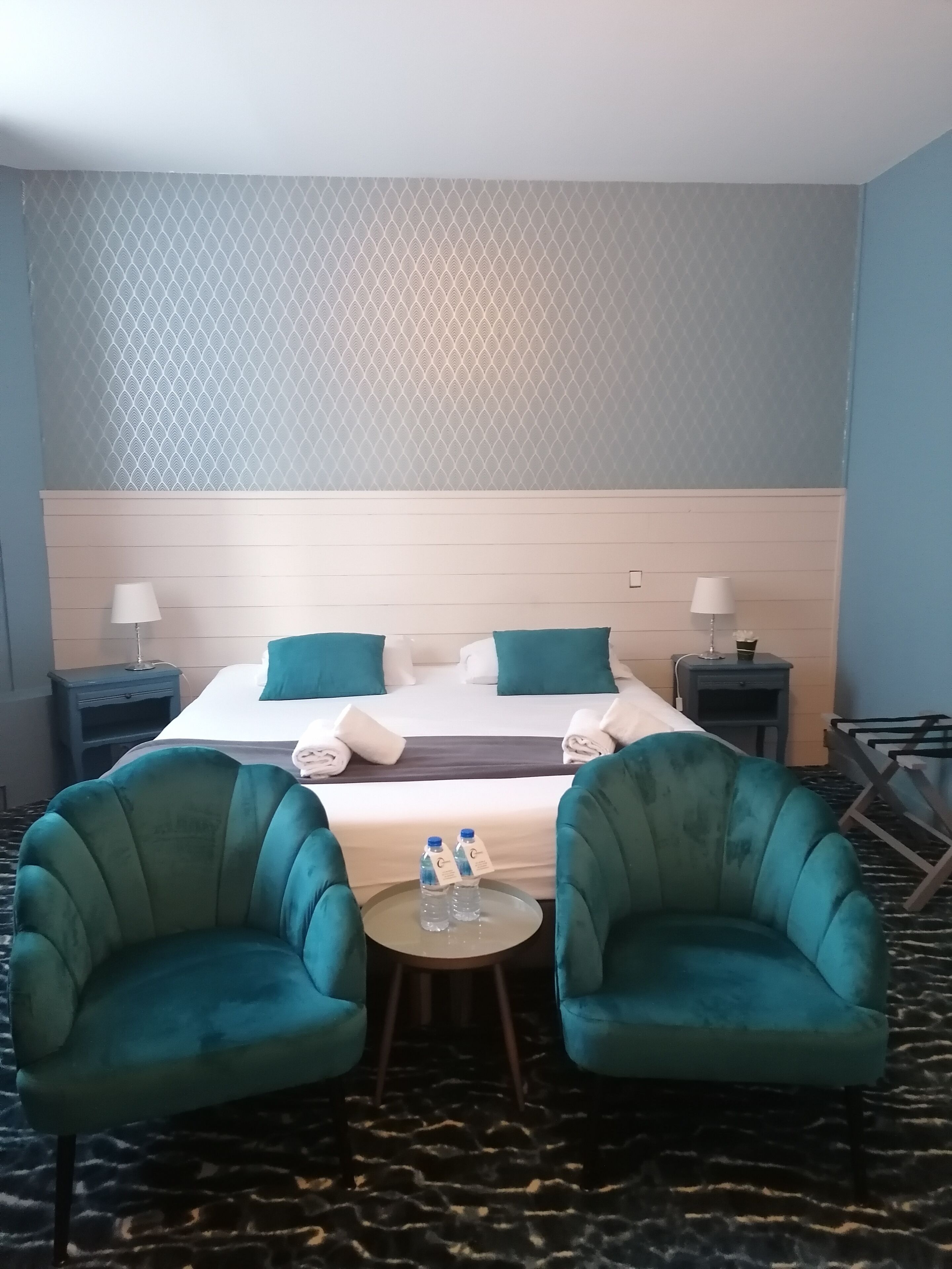 Quarto de hotel com cama, duas cadeiras, uma mesinha com garrafas de água e um tapete estampado.