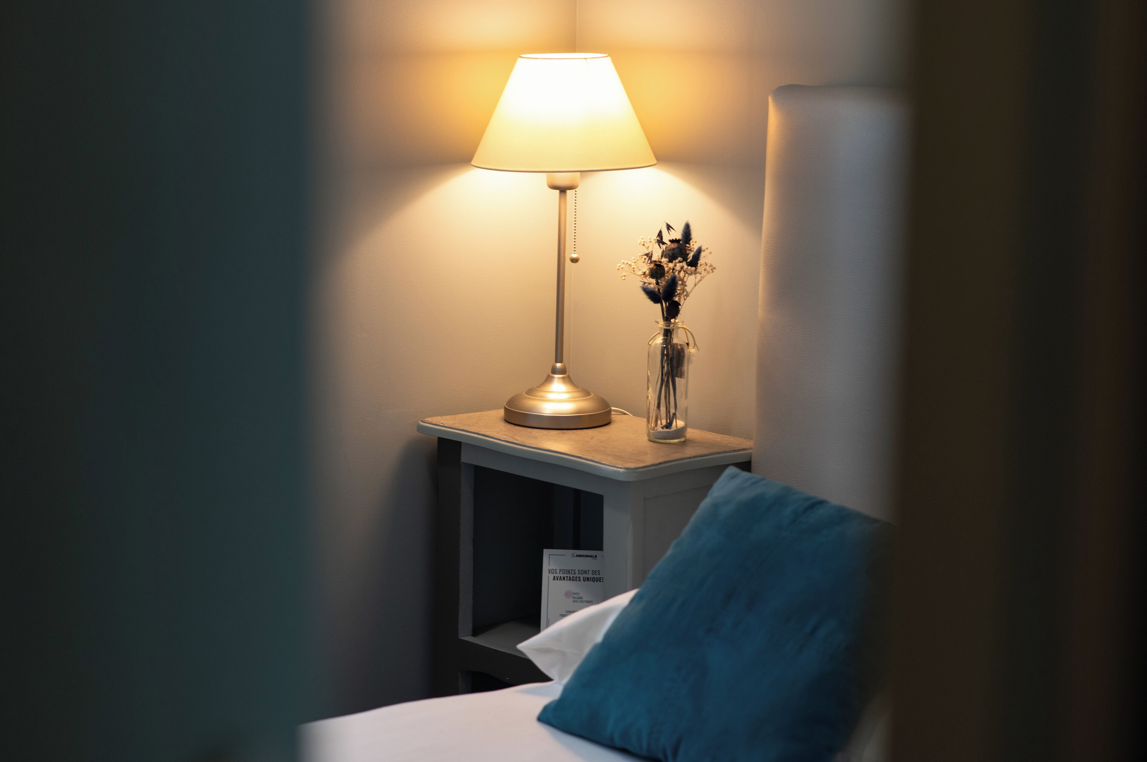 Uma cama bem arrumada com um travesseiro azul, uma mesa de cabeceira com uma luminária e um vaso de flores, e uma placa indicando os recursos do quarto.