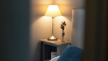 Uma cama bem arrumada com um travesseiro azul, uma mesa de cabeceira com uma luminária e um vaso de flores, e uma placa indicando os recursos do quarto.