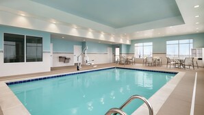 Piscina interna com água azul cristalina, cercada por azulejos bege e uma parede azul claro. Há cadeiras e mesas ao redor da piscina, e uma escada que leva a um nível superior.