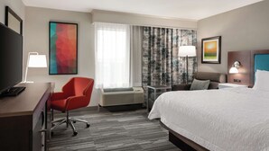 Quarto de hotel com cama, escrivaninha com cadeira, televisão, janela com cortinas e luminária na parede.