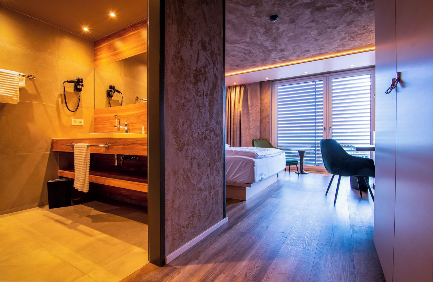 Quarto de hotel moderno com penteadeira de madeira, uma cama, escrivaninha e vista da cidade por grandes janelas.