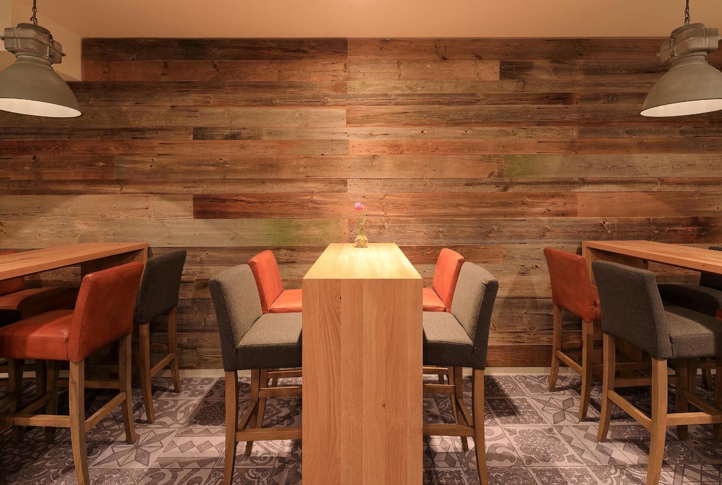 Uma área de refeições com mesas e cadeiras de madeira, uma mesa central com um vaso de flores e uma parede de madeira com luminárias pendentes.