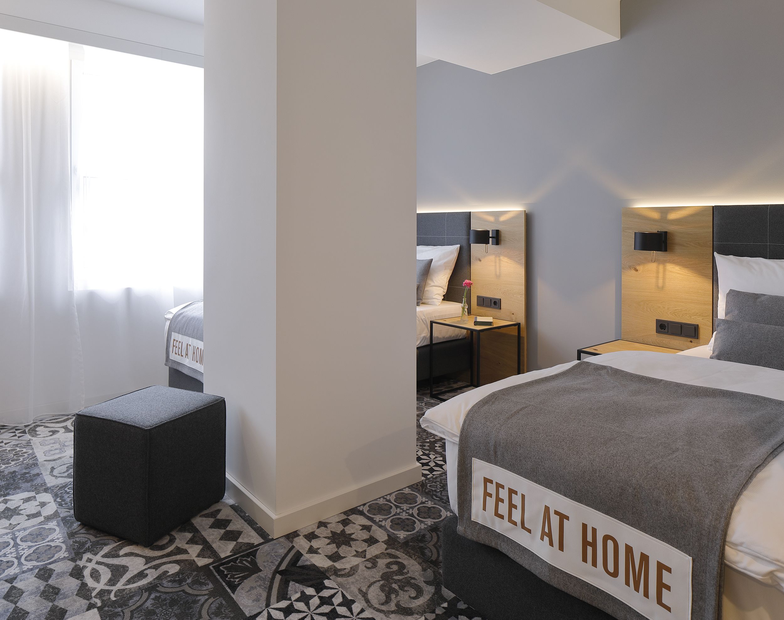 Quarto de hotel com uma cama, mesas de cabeceira, um banquinho pequeno e uma janela com cortinas.