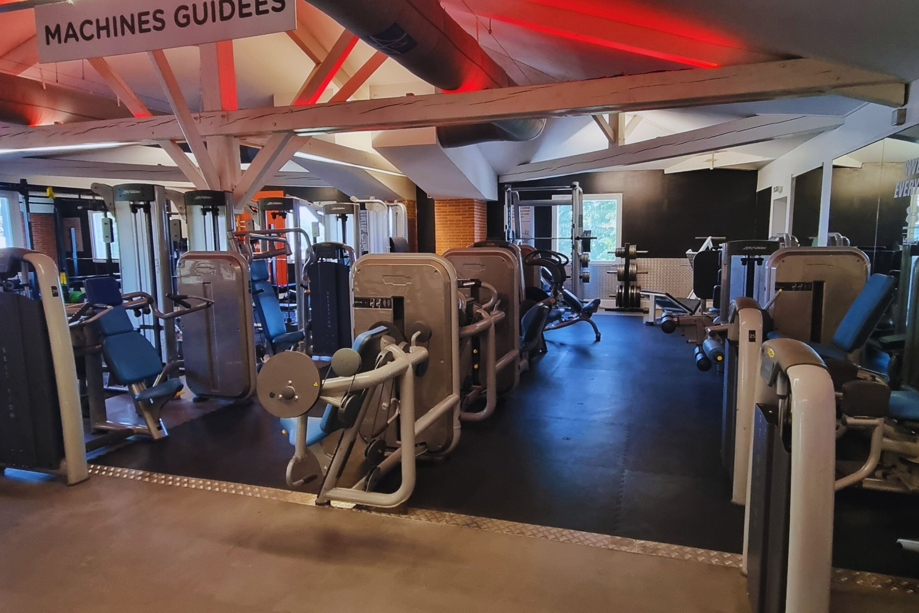 Uma academia com diversos equipamentos de exercício, uma placa com a inscrição 'MACHINES GUIDEES' e uma parede de tijolos ao fundo.