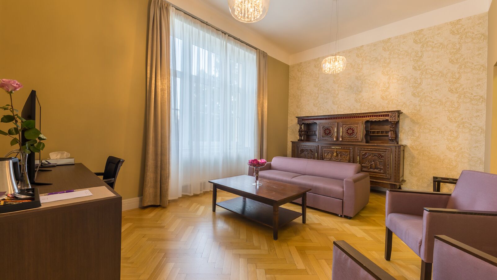 Um quarto com piso de madeira em padrão herringbone, um sofá, uma mesa de centro, uma escrivaninha com um vaso de flores e uma lareira decorada.