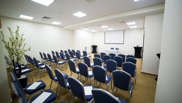 Uma sala de conferências com fileiras de cadeiras azuis, uma tela de projetor e uma lousa.