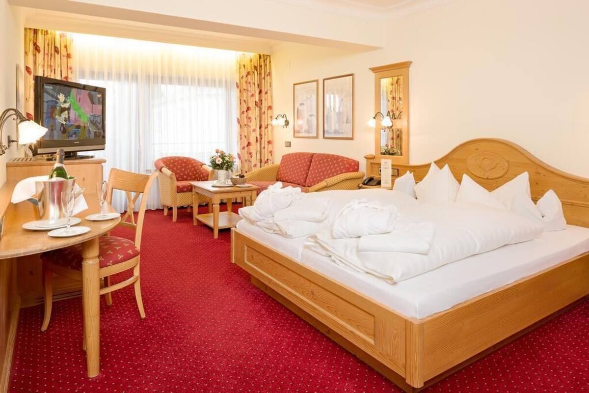 Quarto de hotel com cama de madeira, escrivaninha com televisão, área de refeições e janela com cortinas.