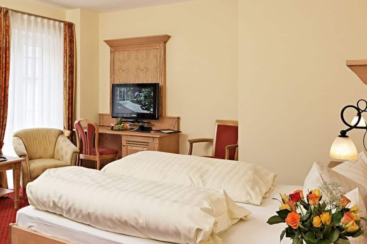 Quarto de hotel com cama, escrivaninha com computador, cadeira, televisão e luminária.