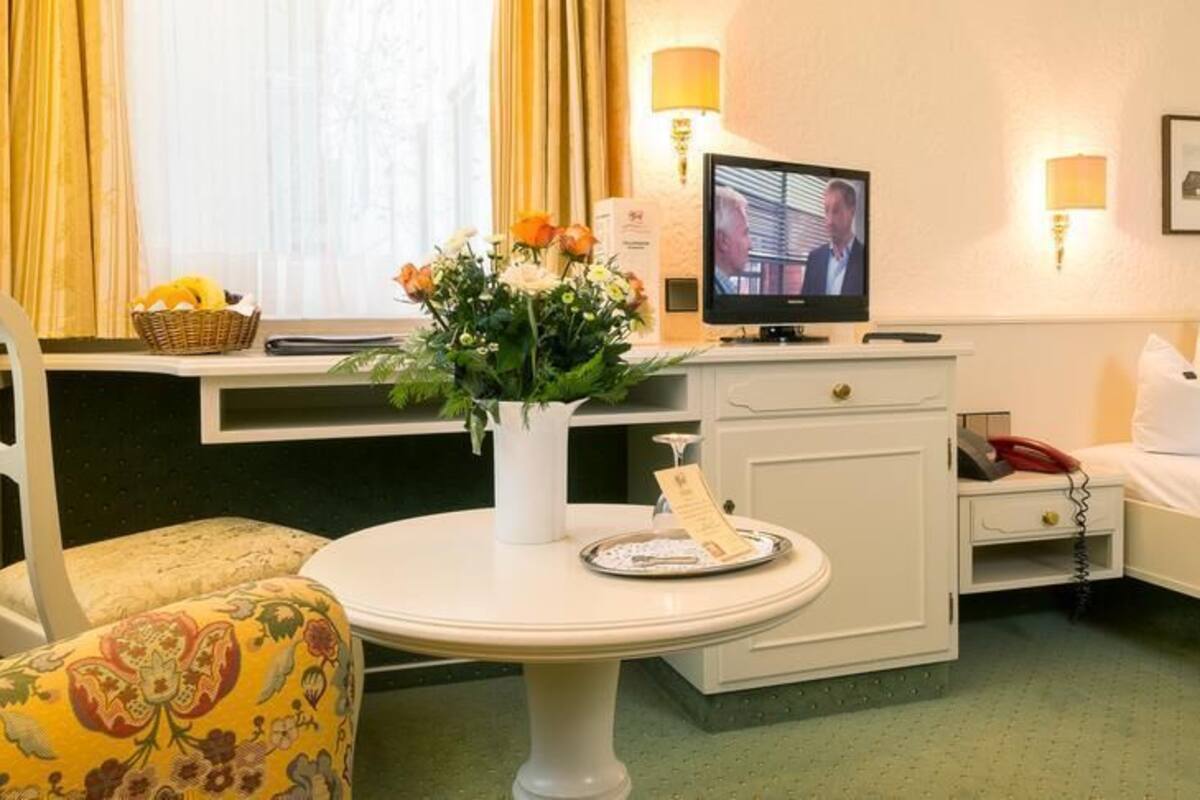 Quarto de hotel com cama, escrivaninha com computador, cadeira, mesinha com um vaso de flores e televisão.