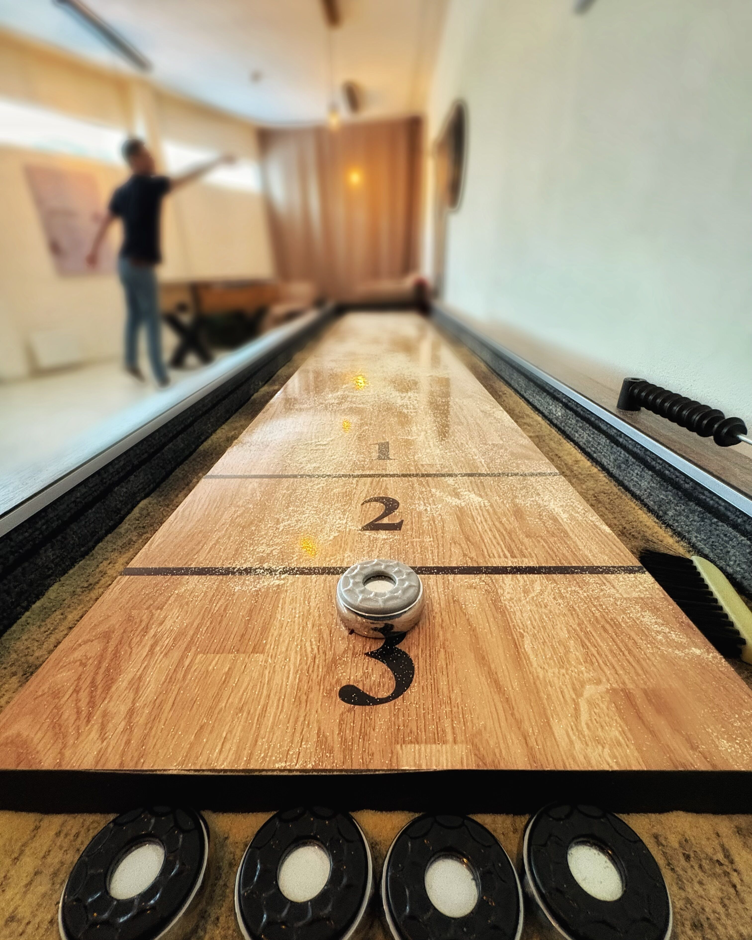 Uma mesa de shuffleboard com áreas de pontuação numeradas e uma pessoa ao fundo.