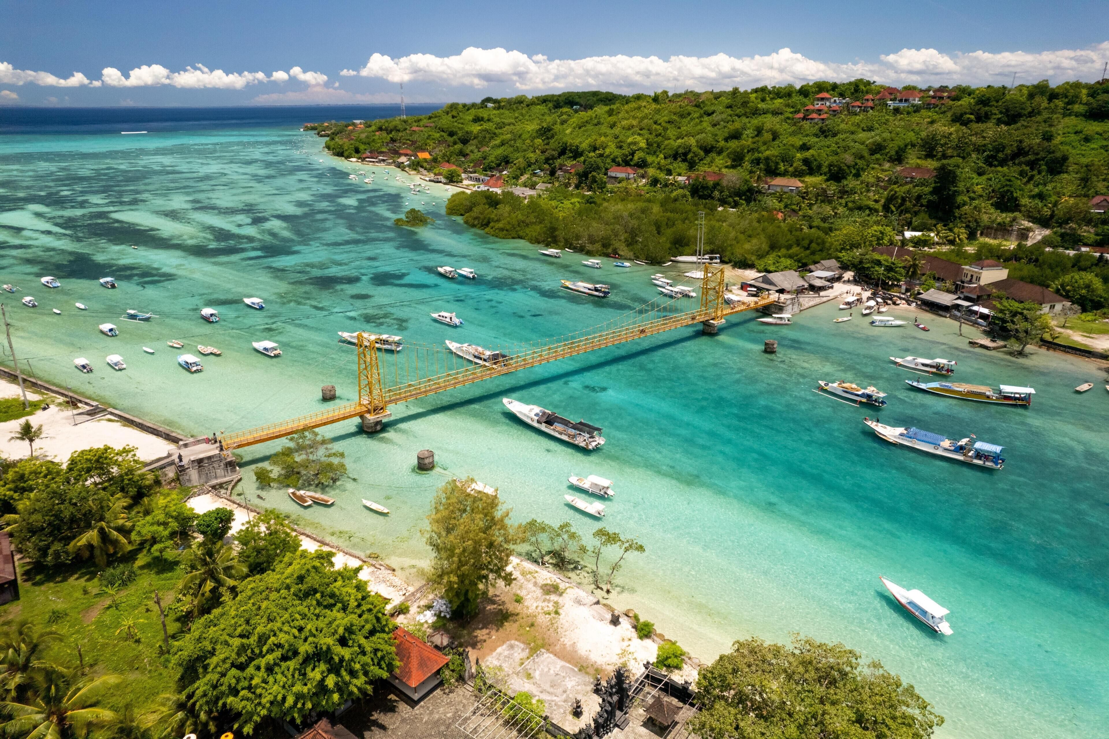 Uma área costeira com uma ponte ligando dois trechos de terra, águas turquesa cristalinas e vários barcos.
