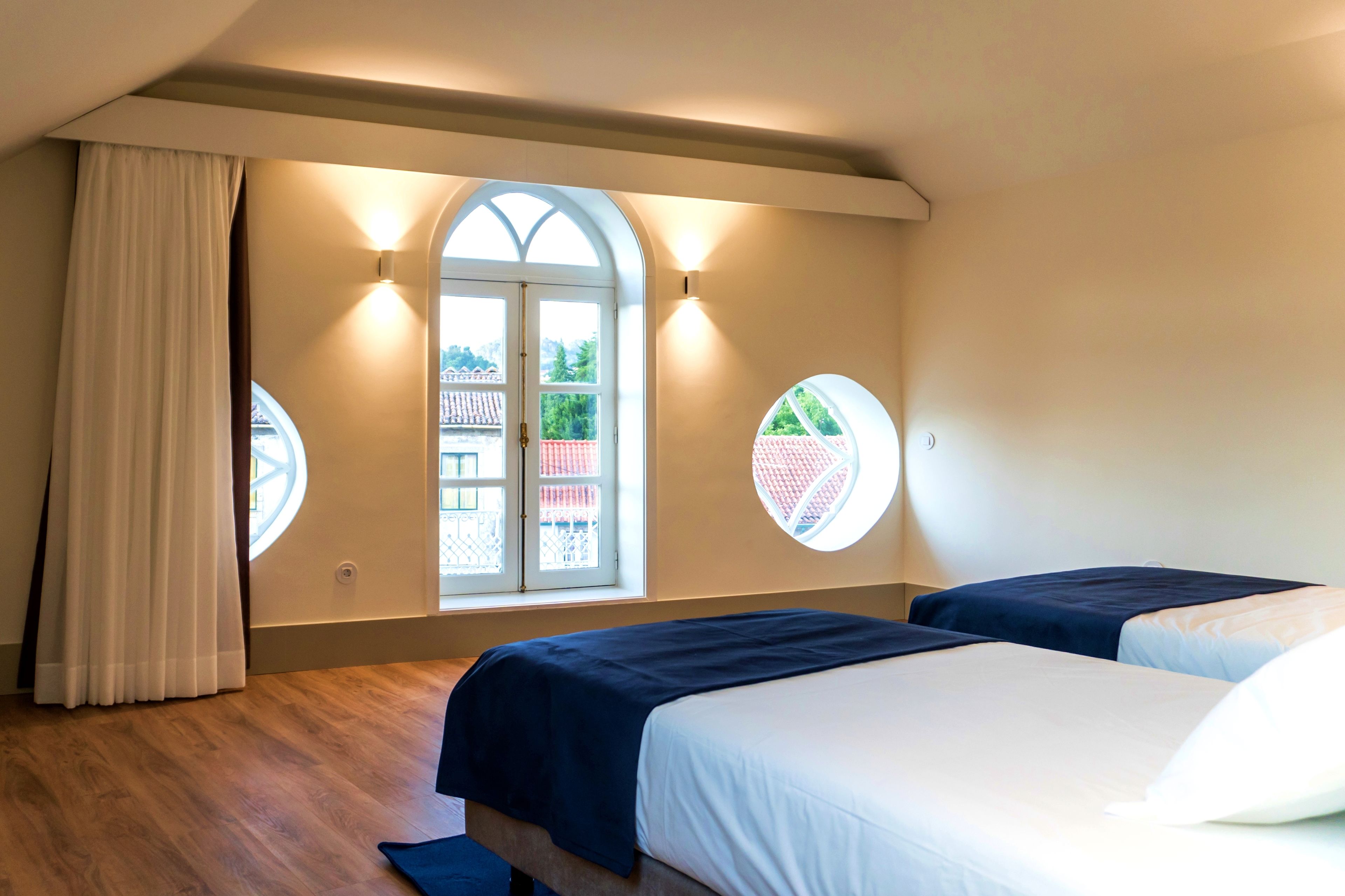Quarto com duas camas, uma grande janela em arco e duas luzes fixadas na parede.