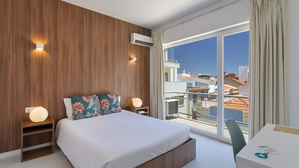 Quarto de hotel moderno com uma cama grande, painéis de madeira, uma escrivaninha e uma varanda com vista para a cidade.
