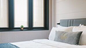 Uma cama bem arrumada com cabeceira cinza, um pequeno vaso de planta no parapeito da janela e uma parede branca com textura discreta.