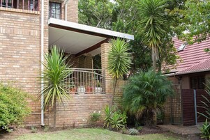 Uma casa de tijolos com varanda coberta, cercada por diversas plantas tropicais.