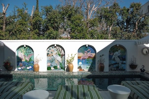 Uma área de piscina com piso de azulejos, um espaço de estar com almofadas listradas e quatro janelas em arco com trabalhos em mosaico.