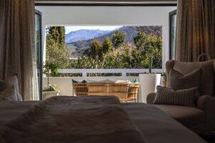 Quarto com cama, uma cadeira, uma mesinha e vista para uma paisagem montanhosa pela janela.
