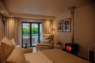 Quarto de hotel com cama, poltrona, mesa com garrafa e copos, lareira e vista para o verde pela janela.