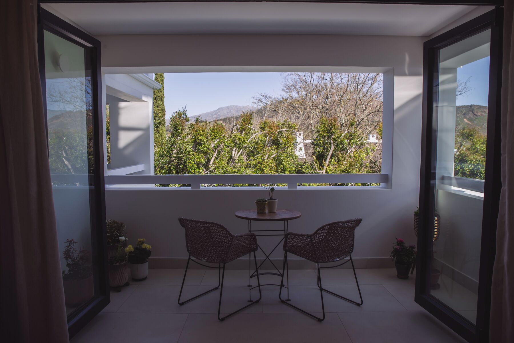 Uma varanda com mesa e duas cadeiras, vista para árvores e colinas, e plantas em vasos.