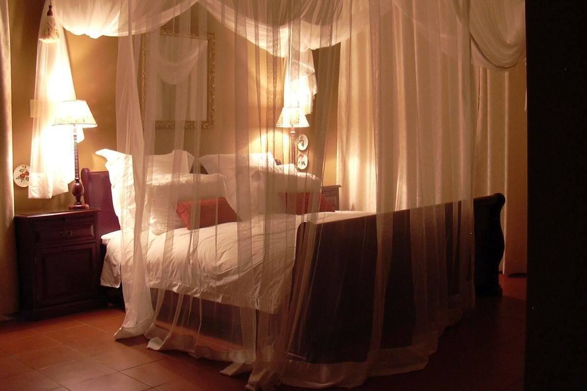 Quarto com cama coberta por um dossel fino, duas luminárias de cabeceira, um criado-mudo e um relógio fixado na parede.