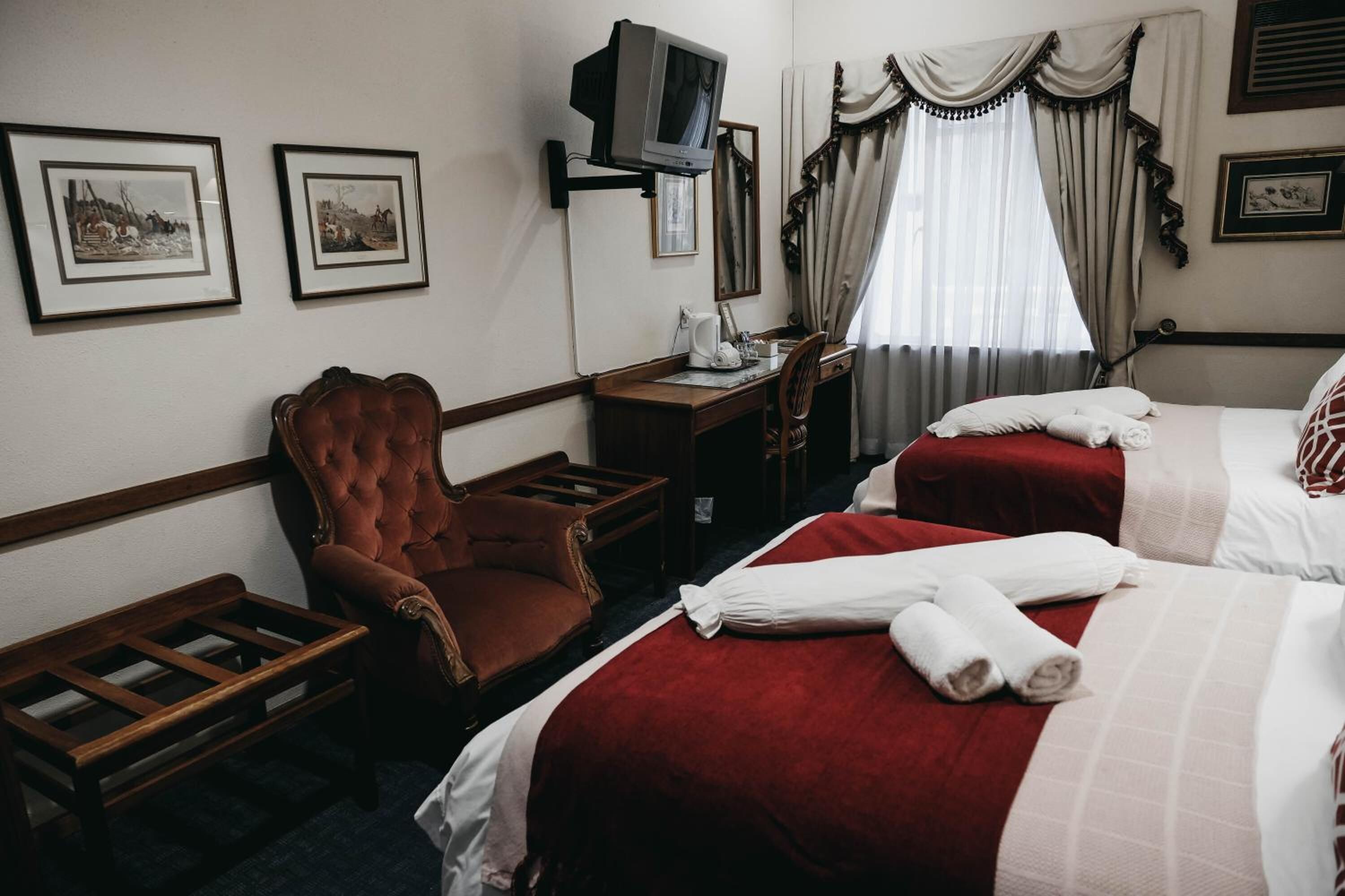 Quarto de hotel com duas camas, uma cadeira de madeira, uma mesa com televisão e quadros nas paredes.