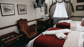 Quarto de hotel com duas camas, uma cadeira de madeira, uma mesa com televisão e quadros nas paredes.