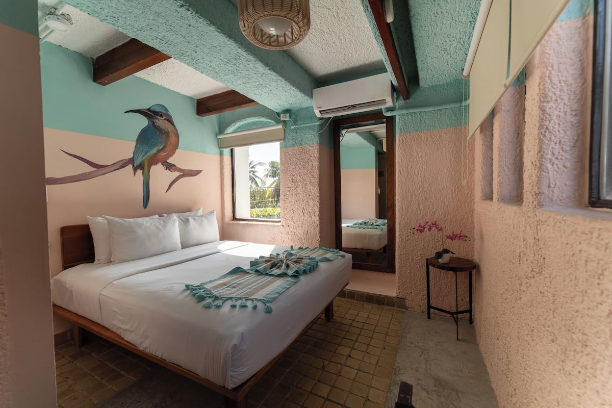 Quarto com cama, painel decorativo de um pássaro, janela e uma pequena mesa com flores.