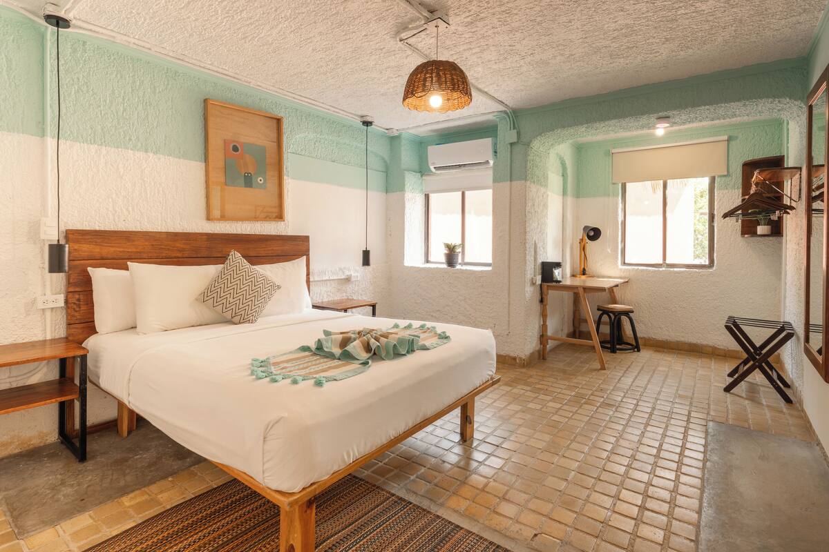 Quarto com cama de madeira, uma secretária pequena, uma cadeira e janela com vista.